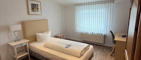 Quarto com cama, mesa de cabeceira, luminária, quadro na parede, janela com cortinas, radiador e escrivaninha com cadeira.