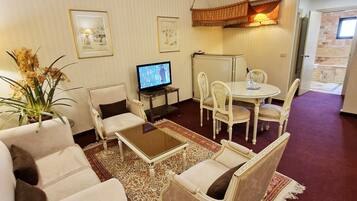 Uma sala de estar com sofá, poltronas, mesa de centro, televisão, mesa de jantar com cadeiras, tapete estampado e luminária pendente decorativa.