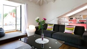 Uma sala de estar moderna com um sofá preto, uma mesa de centro de vidro e um vaso com flores. Há uma janela com vista para um edifício vermelho lá fora.