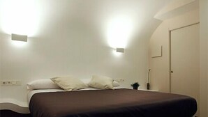 Quarto moderno com cama grande, duas luminárias na parede, mesa de cabeceira pequena e uma porta.