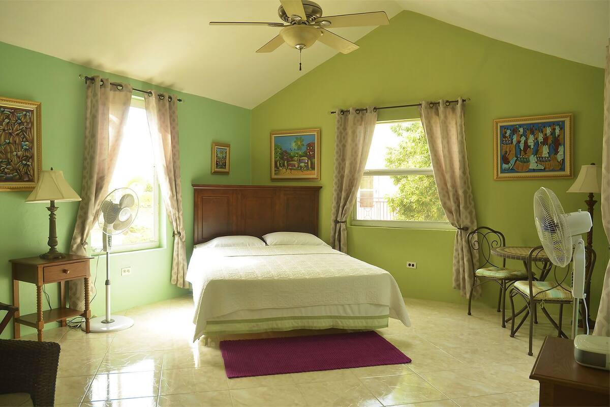 Quarto com cama, ventoinha no teto, escrivaninha com cadeira e duas janelas com cortinas.