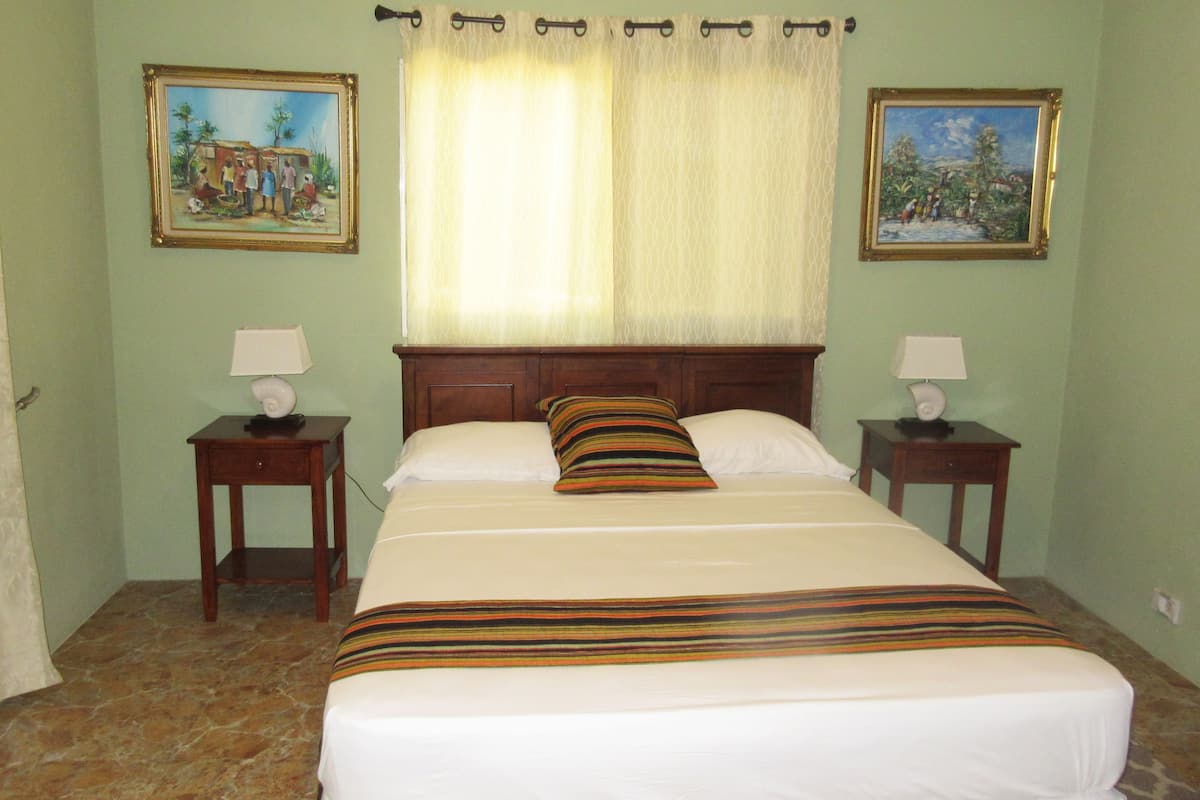 Cama bem arrumada com travesseiro listrado, duas mesas de cabeceira com abajures e dois quadros nas paredes.