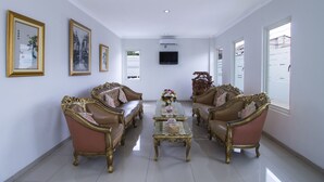 Uma sala de estar espaçosa com mobiliário ornamentado, uma televisão de tela plana na parede e grandes janelas que proporcionam iluminação natural.