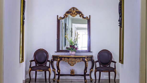 Um quarto com um espelho grande e ricamente decorado, duas poltronas escuras e um pequeno mesa com um vaso de flores.