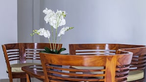 Uma área de refeições com cadeiras de madeira e uma mesa, uma planta em vaso com flores brancas, e uma parede lisa ao fundo.