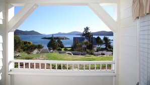 Vista de um alpendre com guarda-corpo branco, com vista para um corpo d'água, montanhas e uma área gramada com árvores e uma cerca.
