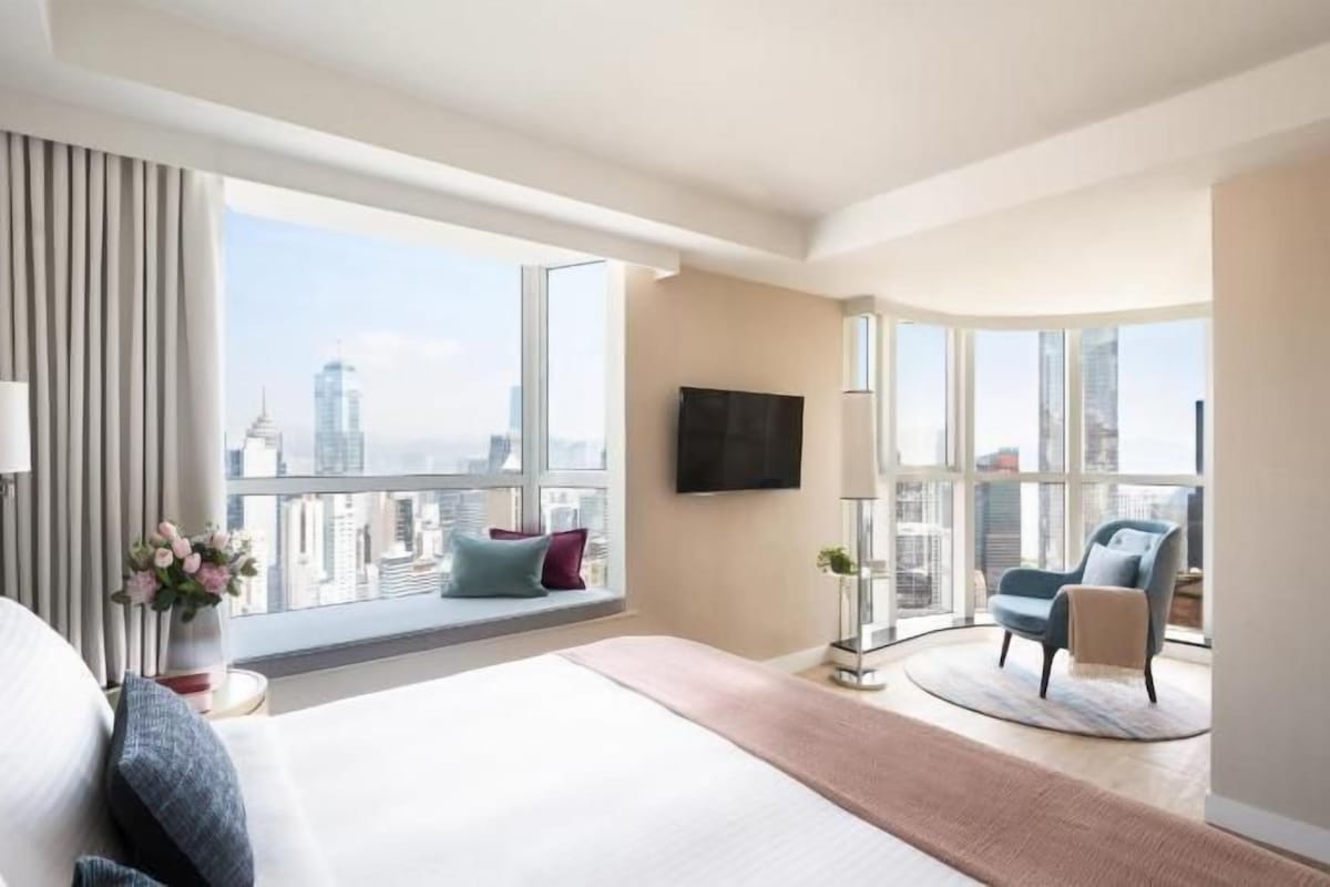 Quarto de hotel com uma cama grande, área de estar com uma poltrona, televisão na parede e janela com vista para a cidade.