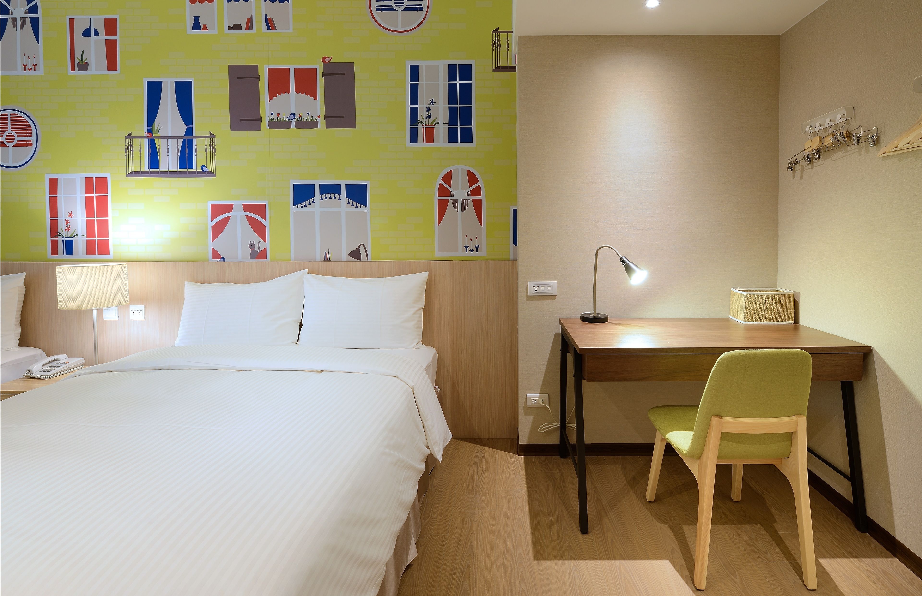 Quarto de hotel com cama, escrivaninha, cadeira, abajur e parede com desenhos coloridos nas janelas.