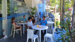 Um café ao ar livre com mesas e cadeiras, rodeado por vegetação e uma estrutura de teto azul.