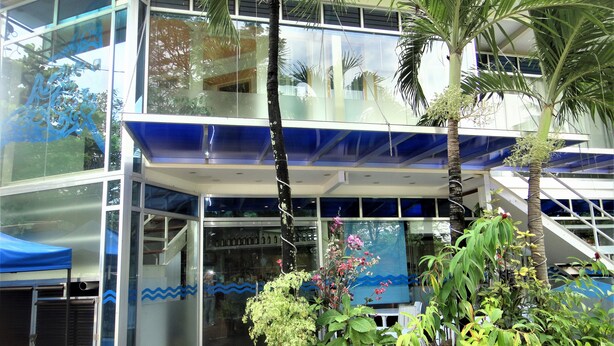 Um edifício moderno com amplas janelas de vidro, entrada coberta e diversos vasos de plantas.