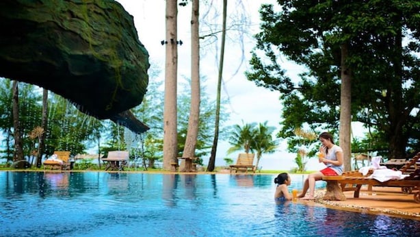 Área de piscina com formação rochosa natural acima, cercada por árvores altas e cadeiras de descanso.