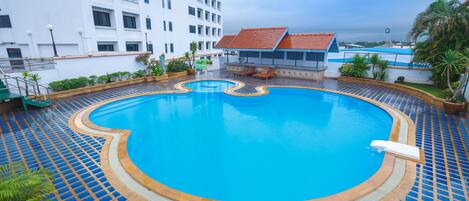 Piscina com deck revestido em azulejos, cercada por um edifício com telhado em telhas vermelhas e fachada branca.