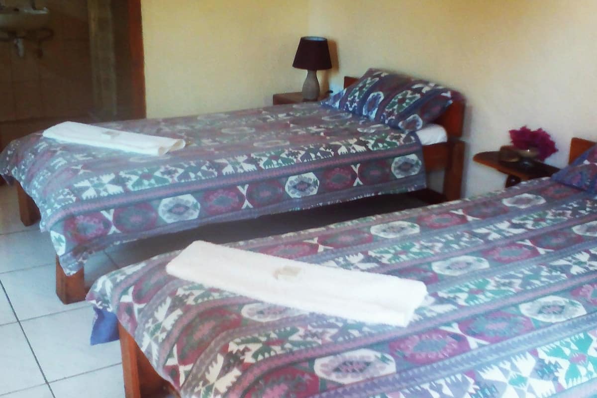 Quarto de hotel com duas camas, cada uma com roupa de cama estampada e uma toalha branca. Há uma mesa de cabeceira com uma luminária e uma pequena mesa com um vaso de flores.