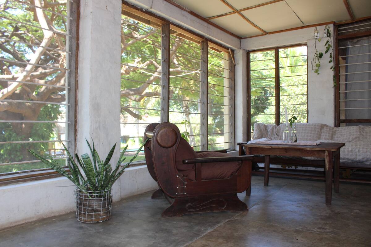 Quarto com grandes janelas, uma cadeira de madeira, uma mesa e um vaso com planta.