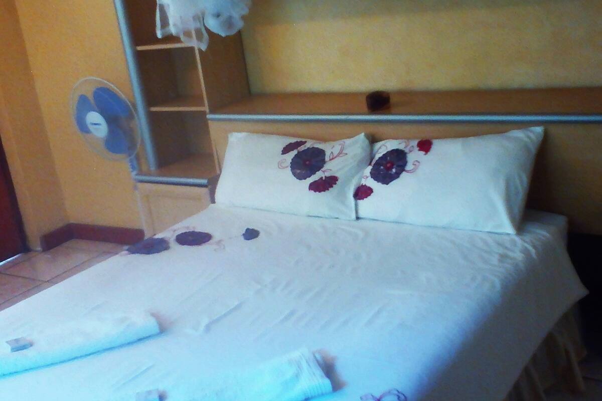 Cama com roupa de cama branca, almofadas decorativas, cabeceira de madeira e uma pequena prateleira com um ventilador e um bicho de pelúcia.