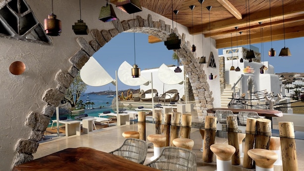 Uma área moderna de refeições ao ar livre com arcos de pedra, móveis de madeira e luminárias pendentes, com vista para o mar e edifícios.