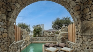 Área de piscina com paredes de pedra e vista através de um arco, com espaço de descanso e almofadas, e uma piscina pequena.