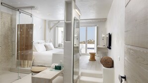 Quarto de hotel moderno com uma cama grande, box de vidro para o chuveiro e vista para a paisagem.