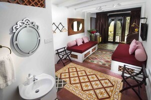 Quarto de hotel com parede branca, espelho redondo, sofá vermelho, cadeira de madeira, mesa pequena com abajur e tapete estampado.