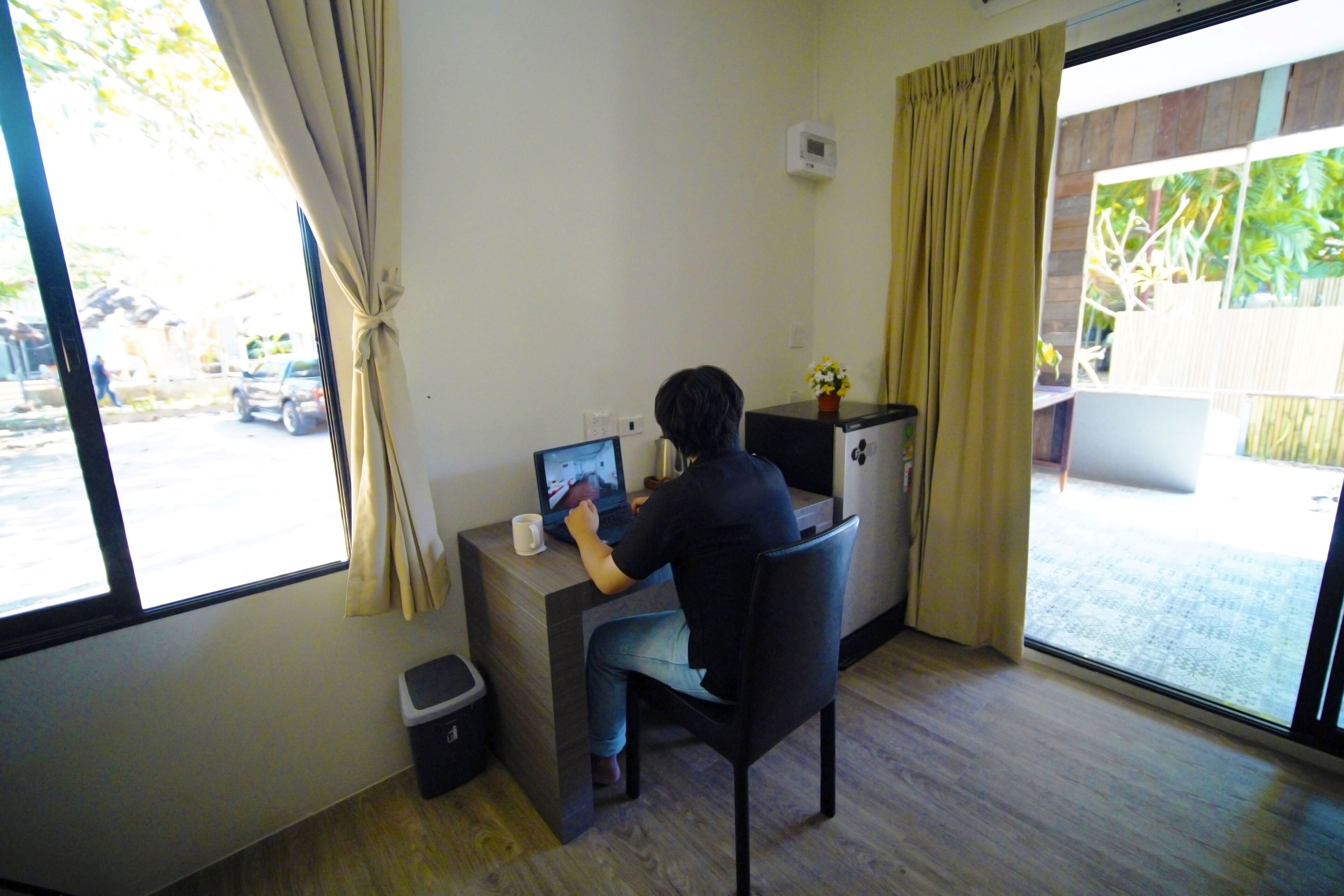 Um quarto com escrivaninha, cadeira, laptop, chávena de café e janela com cortinas.