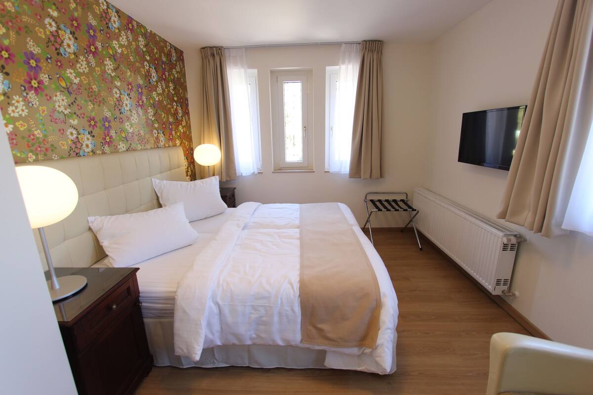 Quarto de hotel com uma cama, televisão, radiador e janela com cortinas.
