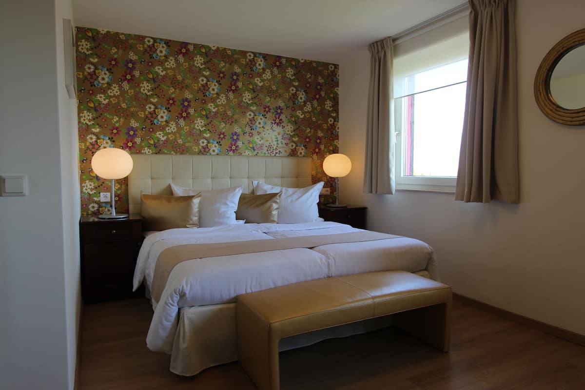 Quarto com uma cama grande, duas mesas de cabeceira, um banquinho, cabeceira com padrão floral e uma janela com cortinas.