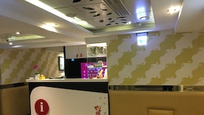 Uma área de recepção com design moderno, destacando um balcão de atendimento com o logotipo 'i' em destaque, uma parede com padrão e um espaço de estar com um sofá bege.