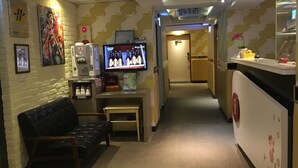 Um saguão de hotel com um sofá, uma máquina de café, uma televisão exibindo garrafas de cerveja e um balcão de recepção.
