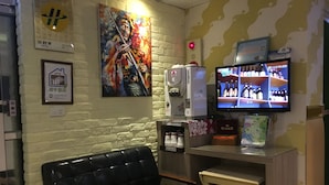 Um saguão de hotel com um sofá de couro preto, uma televisão exibindo uma cena de bar, uma geladeira e uma parede com uma pintura colorida e diversos avisos.