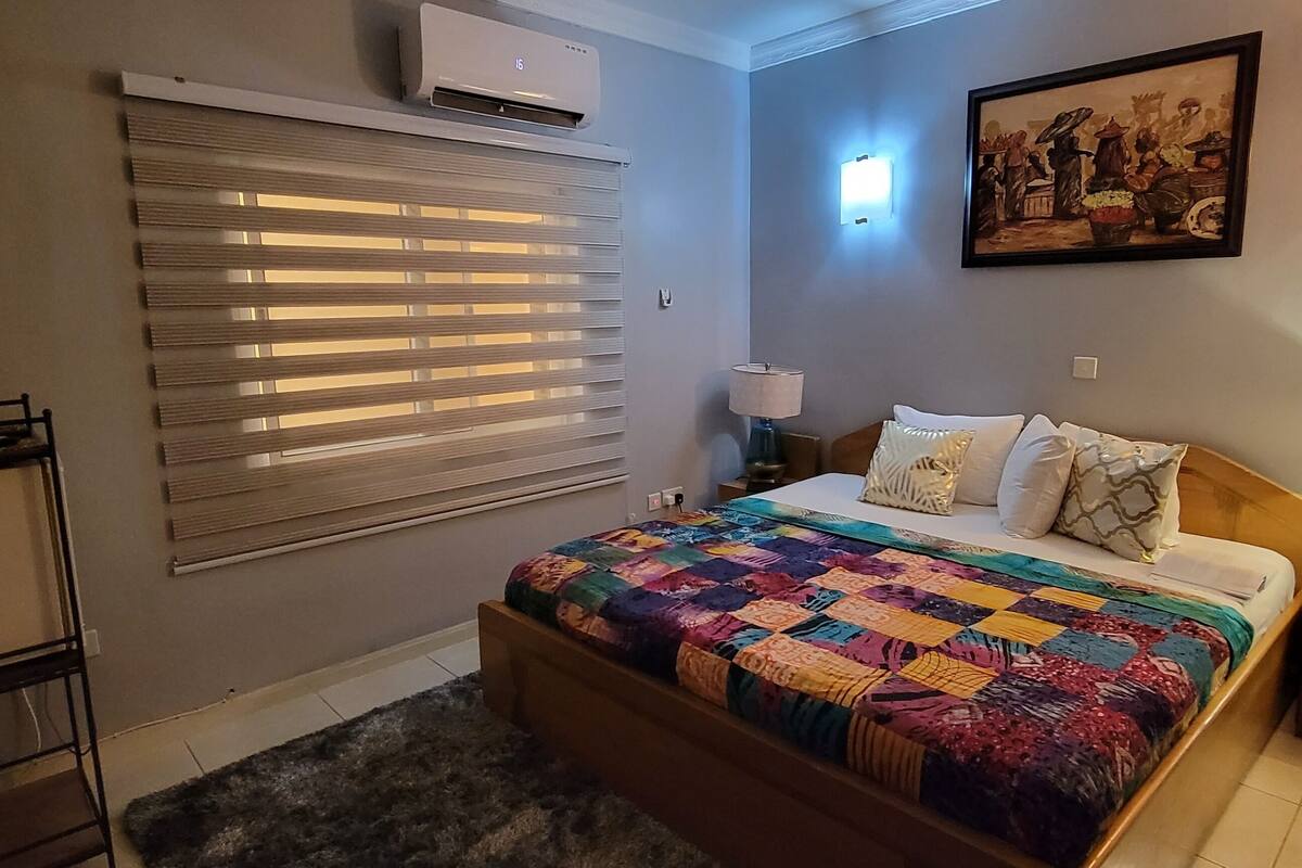 Quarto com cama, janela com persianas, ar-condicionado na parede, mesa de cabeceira com abajur e um quadro na parede.