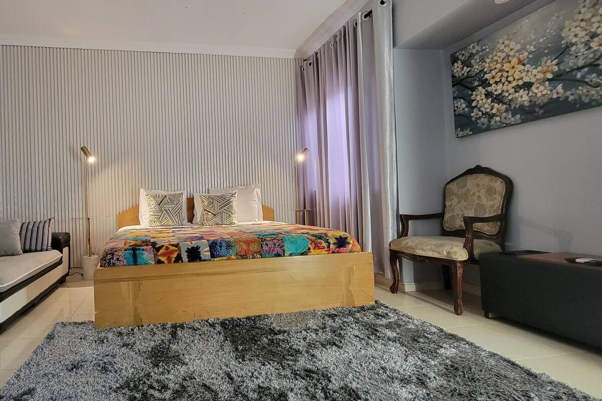 Quarto com cama de madeira, sofá preto, uma cadeira e um quadro grande na parede.
