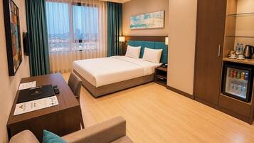 Quarto de hotel com cama, secretária, poltrona, sofá, minibar e vista para a cidade.