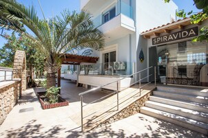 Um edifício moderno com uma placa exibindo 'SPIRAVIDA', uma parede de pedra, uma palmeira e uma escadaria que leva a uma área coberta.