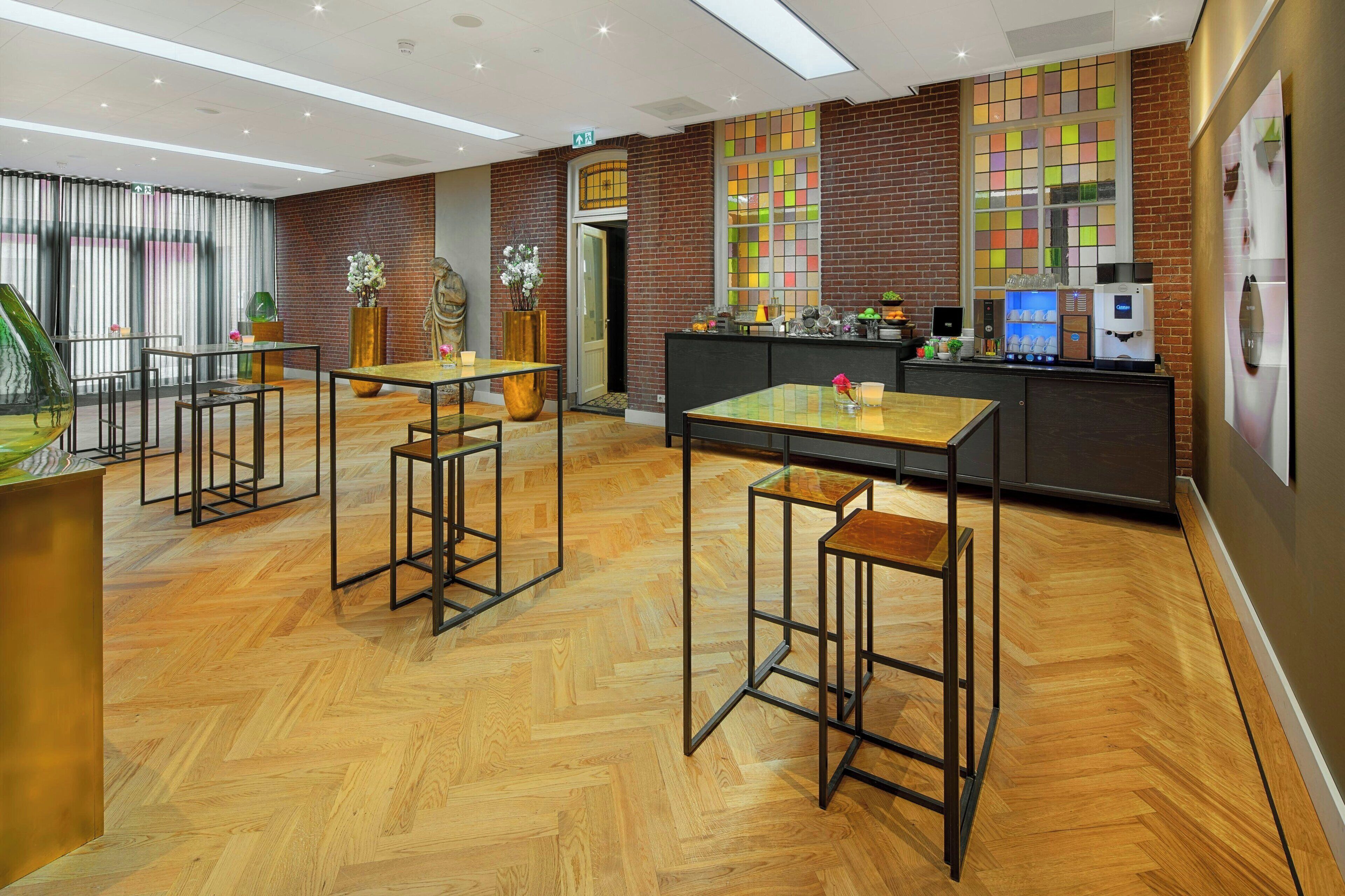Um hall de hotel moderno com piso de madeira, mesas de estrutura metálica e uma parede decorada com tijolos.