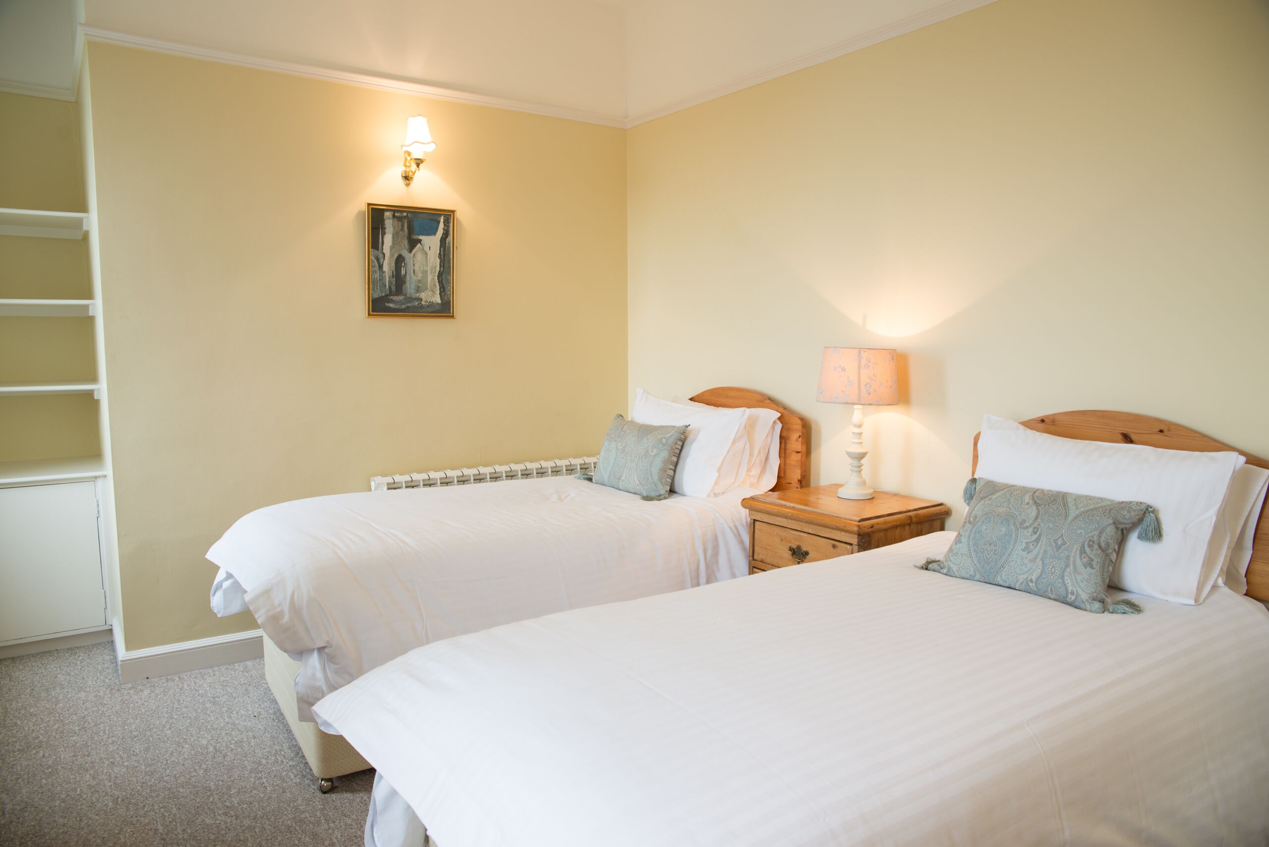 Quarto de hotel com duas camas de solteiro, mesa de cabeceira com abajur, um quadro na parede e uma prateleira embutida.