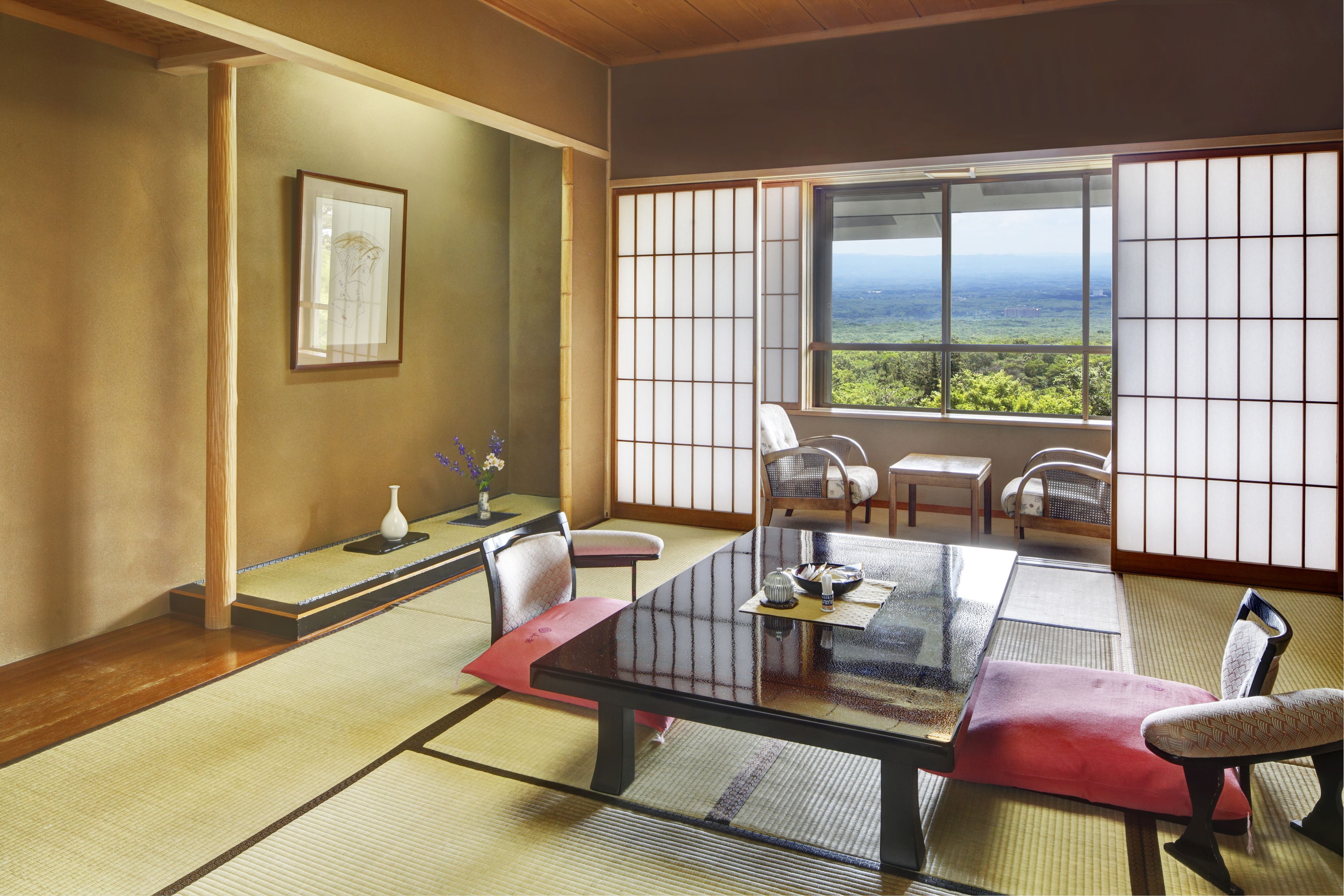 Um espaço com esteiras de tatame, uma mesa baixa, cadeiras, um quadro emoldurado e vista através de grandes biombos shoji.