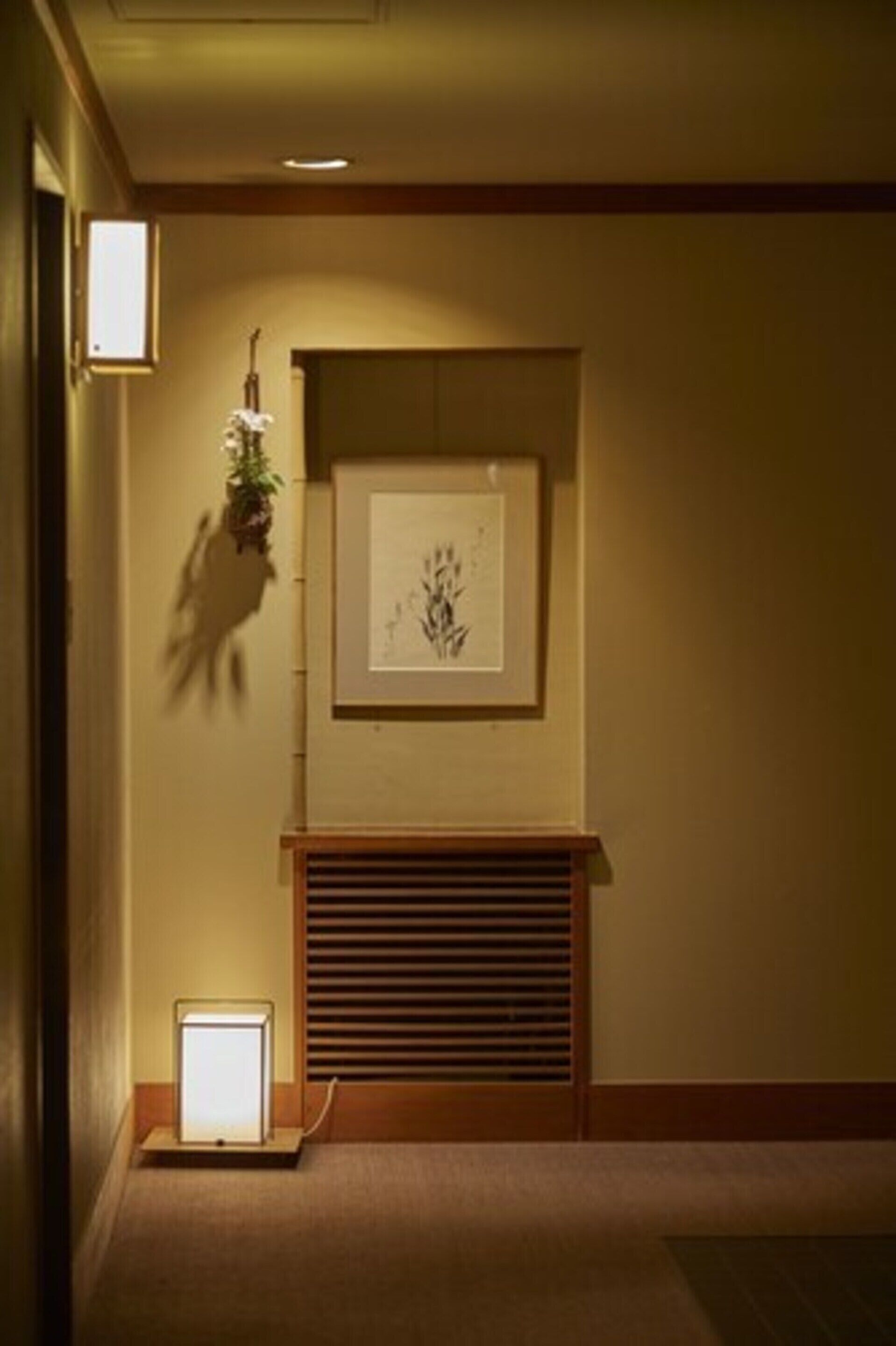 Um corredor com uma obra de arte emoldurada na parede, uma pequena mesa com abajur e um vaso de planta.