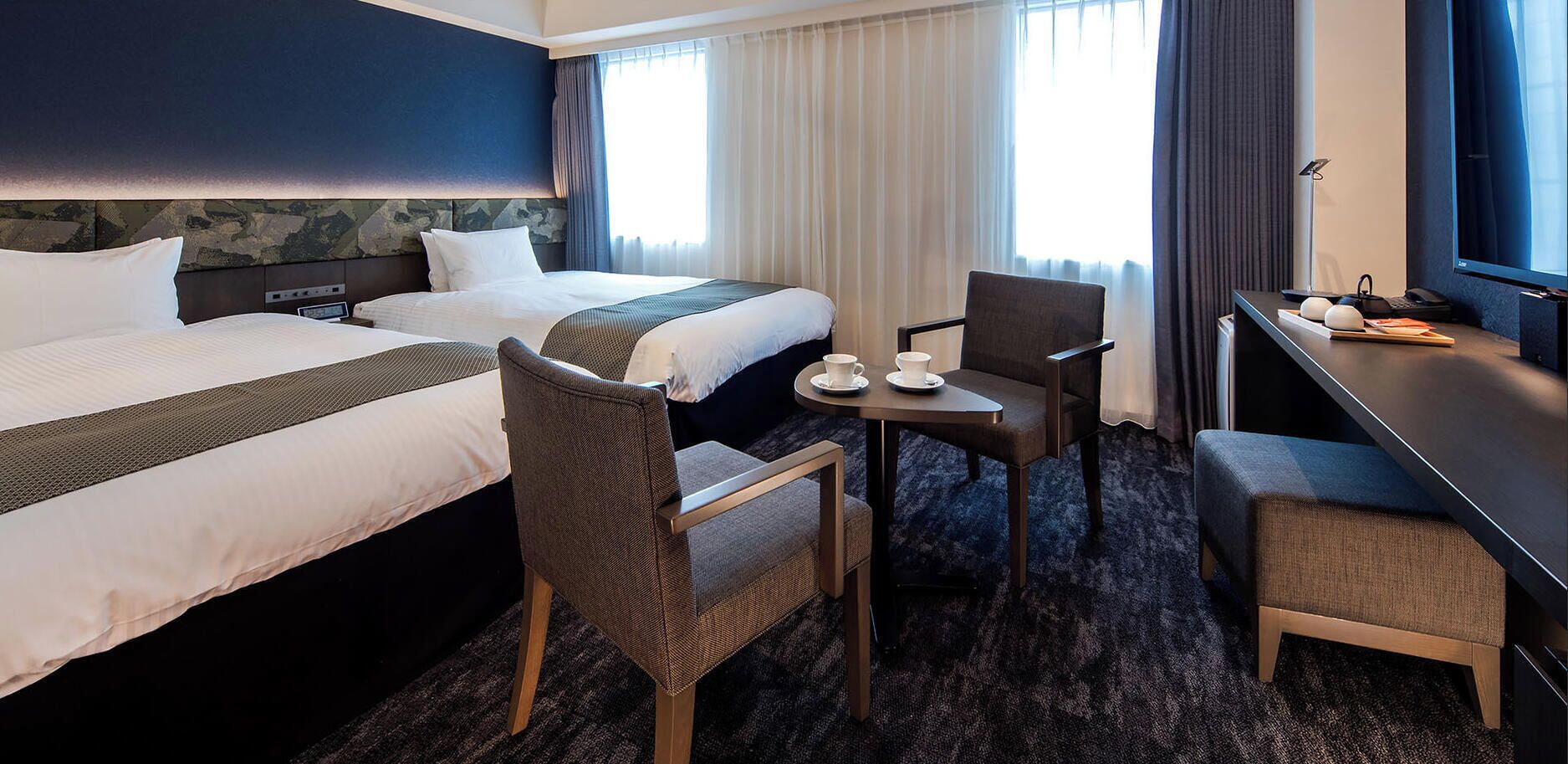 Quarto de hotel com duas camas, uma escrivaninha, uma cadeira e uma pequena mesa com xícara e pires.