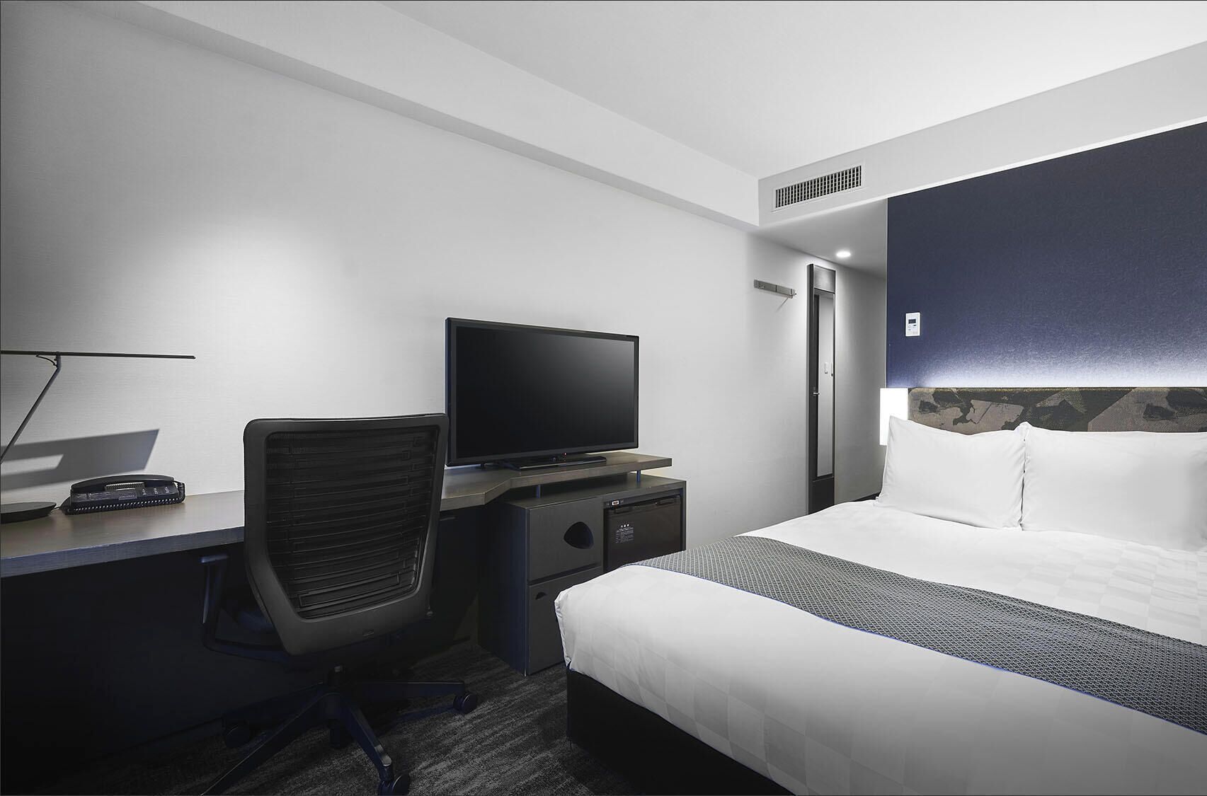 Quarto de hotel com cama, escrivaninha com computador, cadeira, televisão e guarda-roupa.