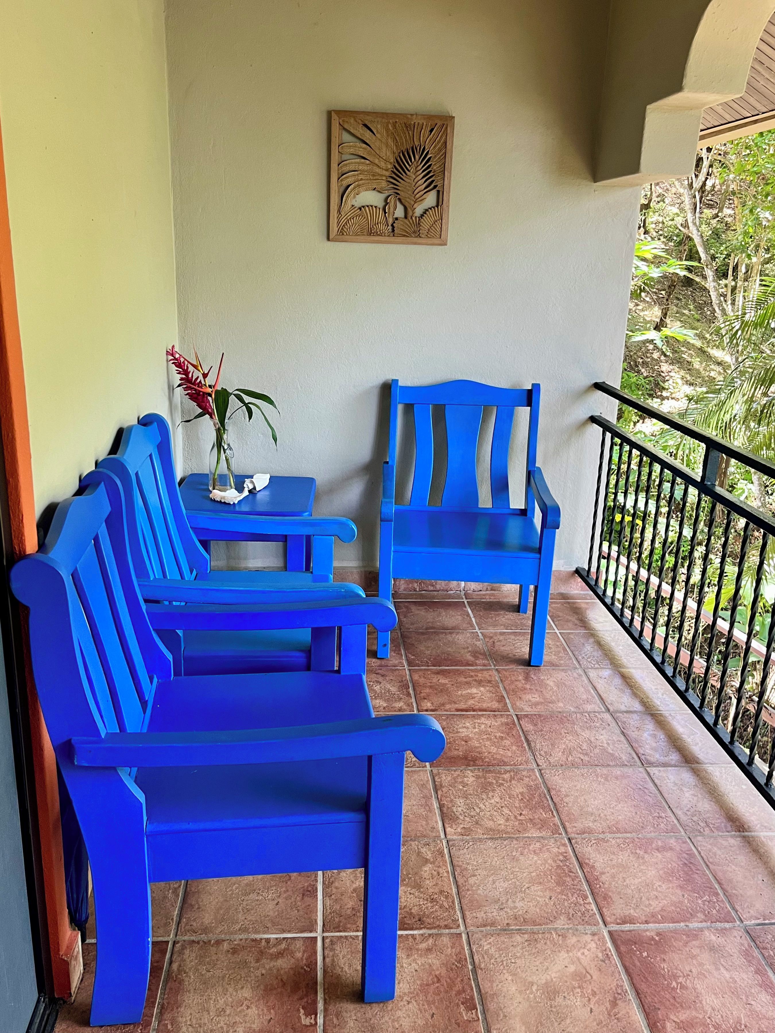 Varanda com cadeiras azuis e uma mesa pequena, um detalhe decorativo na parede e vista para o verde.