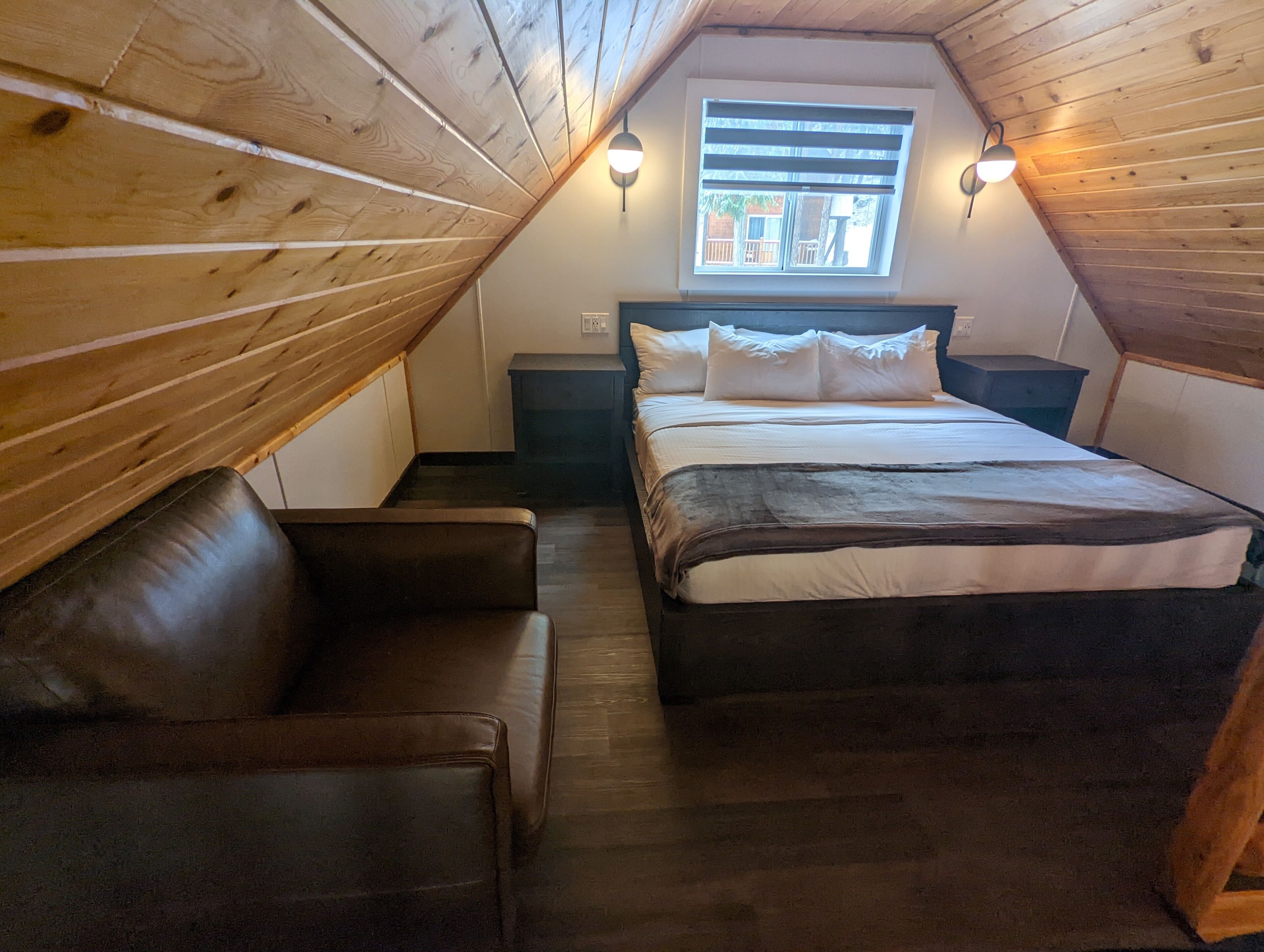 Quarto com teto inclinado de madeira, cama com roupa de cama branca, um banquinho de couro escuro e duas luminárias fixadas na parede.