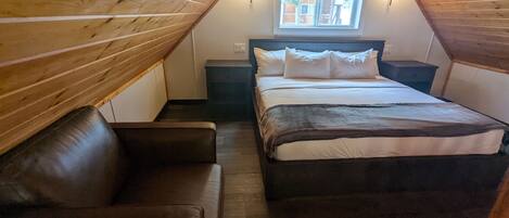 Quarto com teto inclinado de madeira, cama com roupa de cama branca, um banquinho de couro escuro e duas luminárias fixadas na parede.