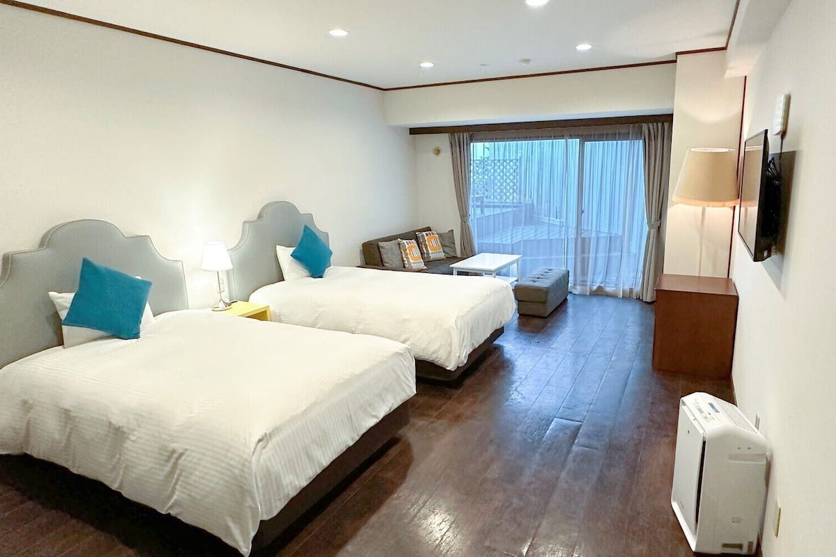 Quarto de hotel com duas camas, um sofá, uma televisão e uma janela com cortinas.