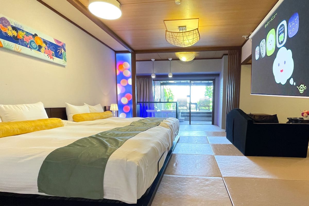 Quarto de hotel com uma cama grande, uma escrivaninha, uma televisão e um projetor.