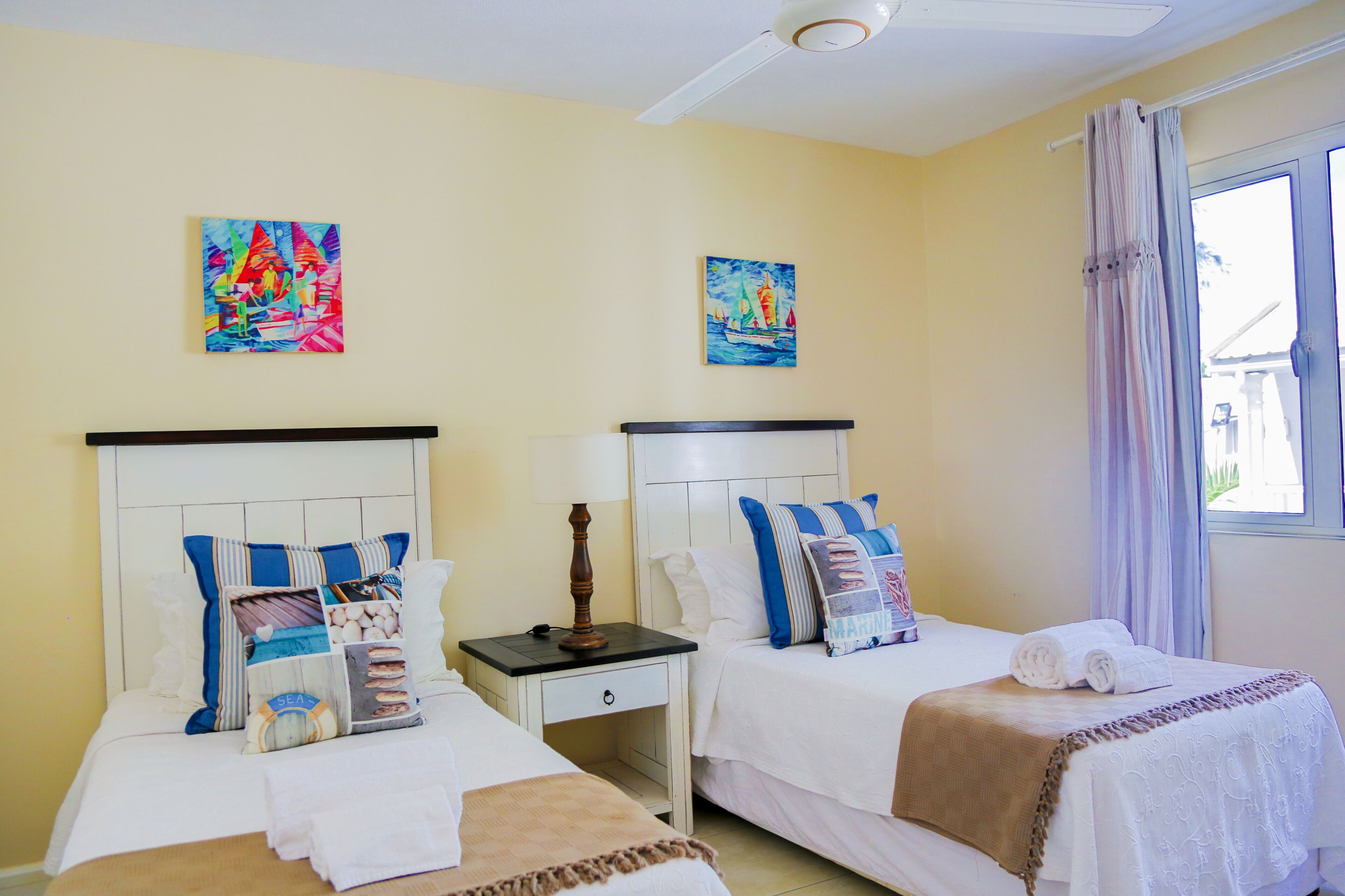 Quarto de hotel com duas camas, cada uma com cabeceira, mesa de cabeceira e abajur. Há quadros nas paredes e uma janela com cortinas.