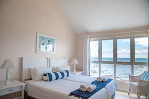 Quarto com cama grande, duas mesas de cabeceira, janela com vista para a praia e uma cadeira.