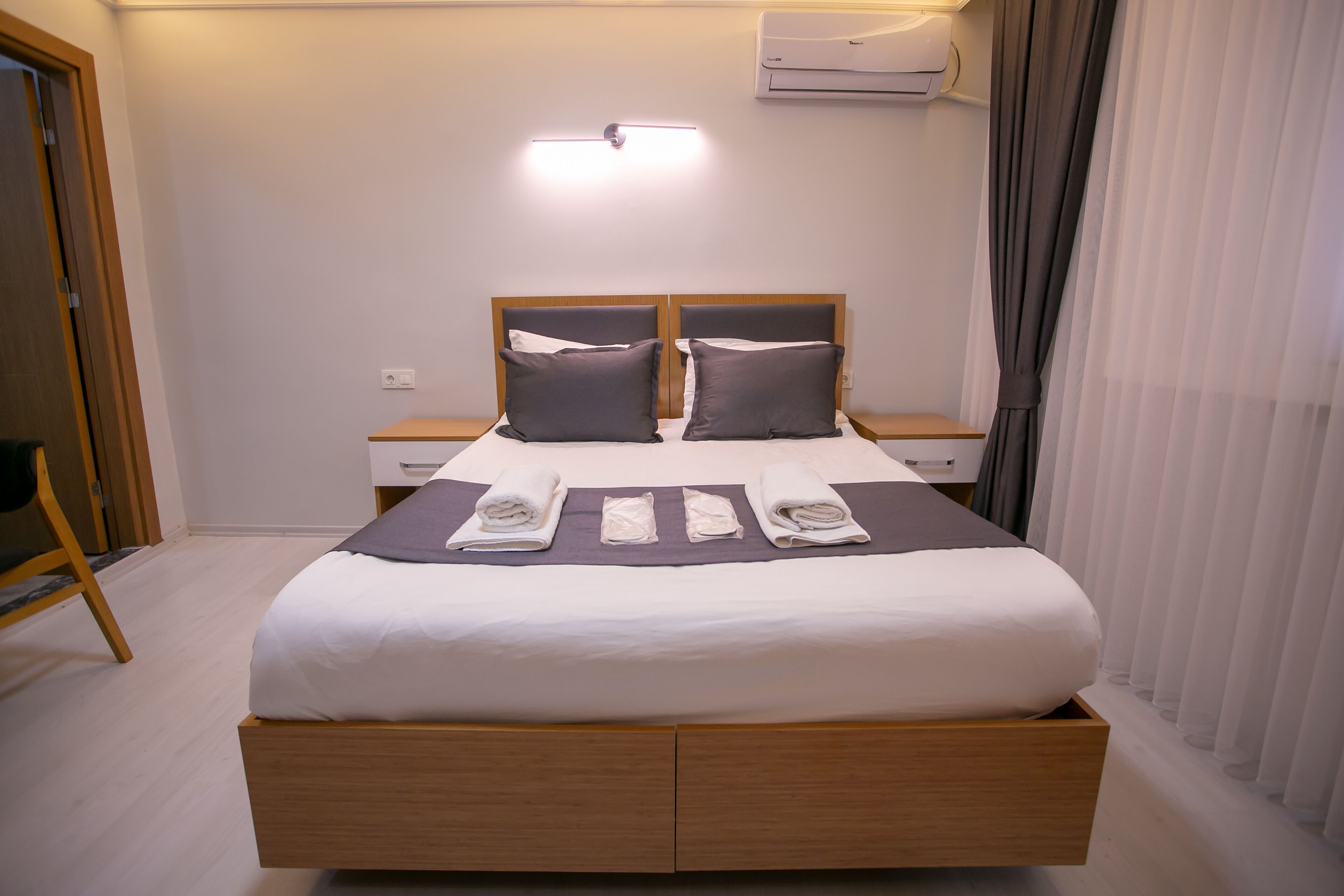 Uma cama bem arrumada com almofadas, uma mesa de cabeceira, um ar-condicionado na parede e uma cortina.