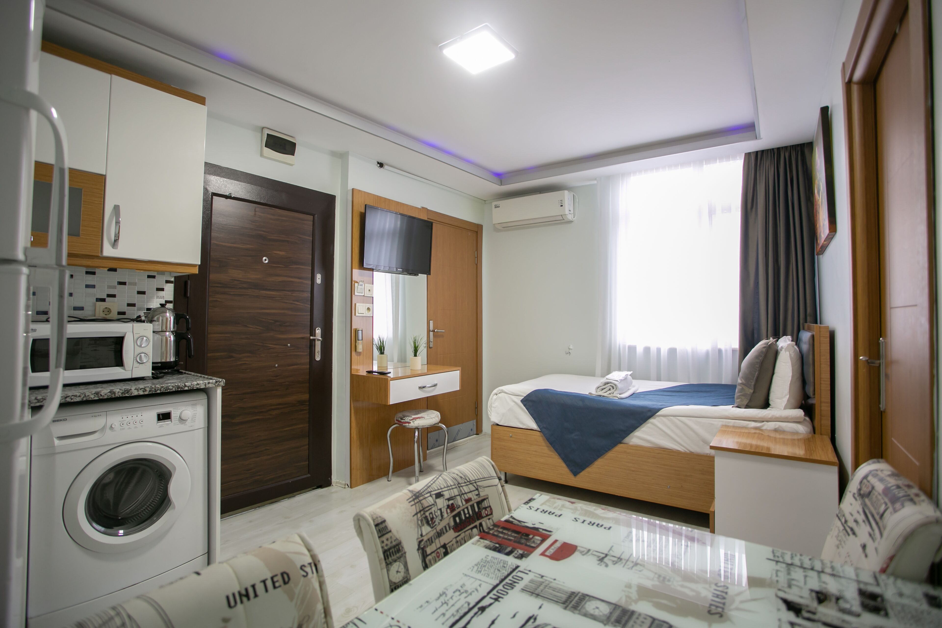 Quarto de hotel compacto com máquina de lavar, micro-ondas, cama com cobertor azul, uma pequena mesa com jornais e TV na parede.