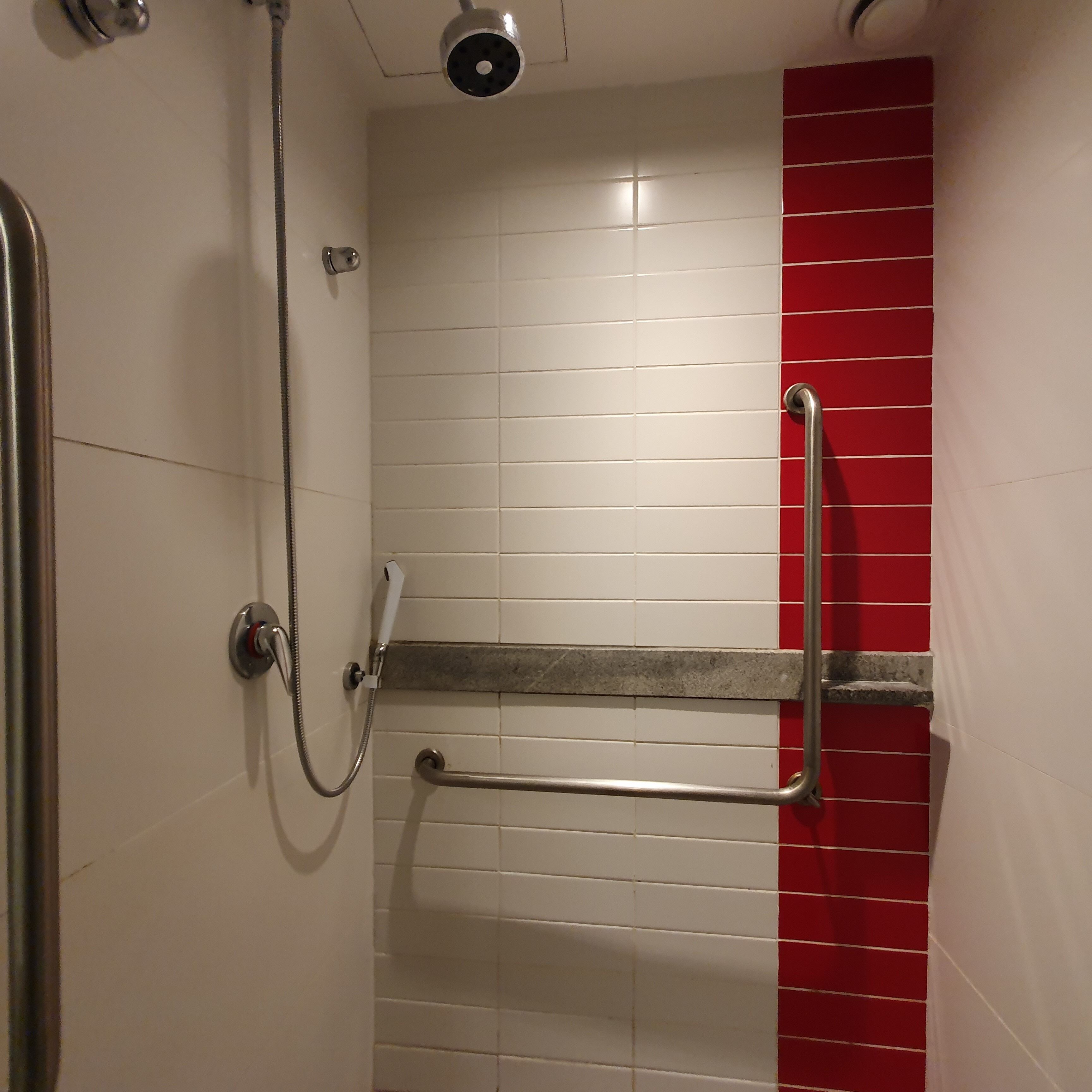 Casa de banho com paredes revestidas a azulejos brancos, uma faixa vermelha, chuveiro com chuveiro manual e barra de apoio.