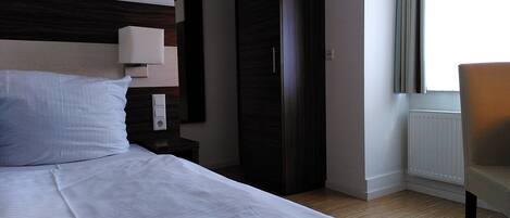 Quarto de hotel com cama bem arrumada, luminária de cabeceira, cadeira e janela com cortinas.